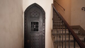 Uma porta de madeira escura com arco pontiagudo e uma janela, que dá acesso a uma escadaria com corrimão de madeira e degraus de pedra.
