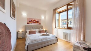 Quarto com cama grande, mesa de cabeceira com abajur, radiador e janela com cortinas.