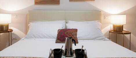 Cama bem arrumada com lençóis brancos, duas luminárias de cabeceira, uma cafeteira e uma bandeja com duas xícaras.