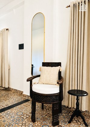 Quarto com espelho grande, cadeira de madeira com almofada, mesa redonda pequena e ladrilhos de chão estampados.