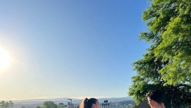 Duas pessoas sentadas a uma mesa ao ar livre, com vista para um horizonte urbano e vegetação ao fundo.