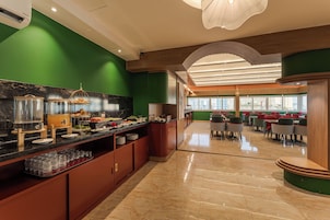 Uma zona de refeições moderna num hotel, com serviço de buffet, área de estar com mesas e cadeiras, e vista da cidade através de grandes janelas.