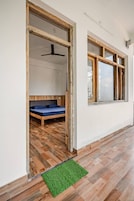 Quarto com piso de madeira, cama com colchão azul, ventoinha no teto e janela com molduras de madeira.