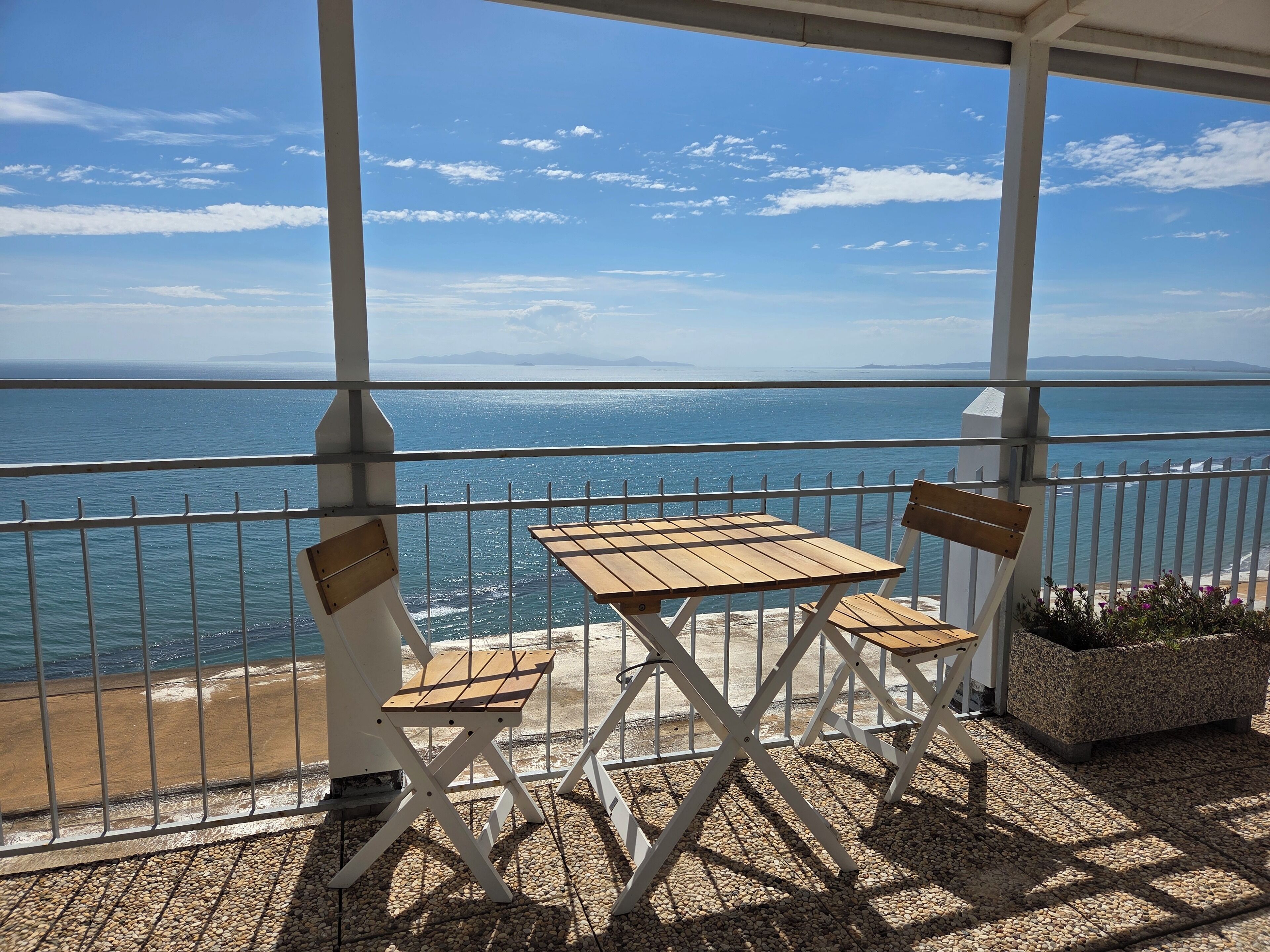 Varanda com mesa e cadeiras de madeira, com vista para uma praia e o mar.