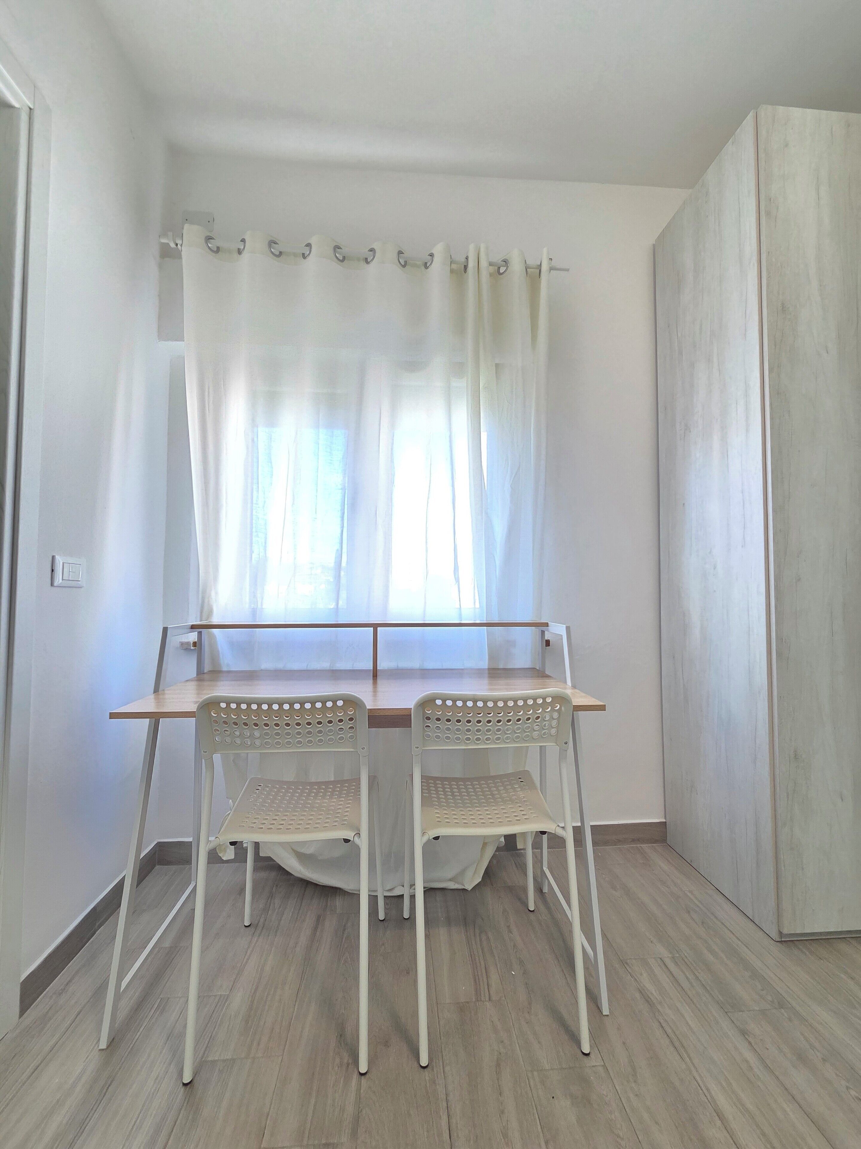 Quarto com mesa de madeira e duas cadeiras brancas, janela com cortinas leves e um guarda-roupa em tom claro.