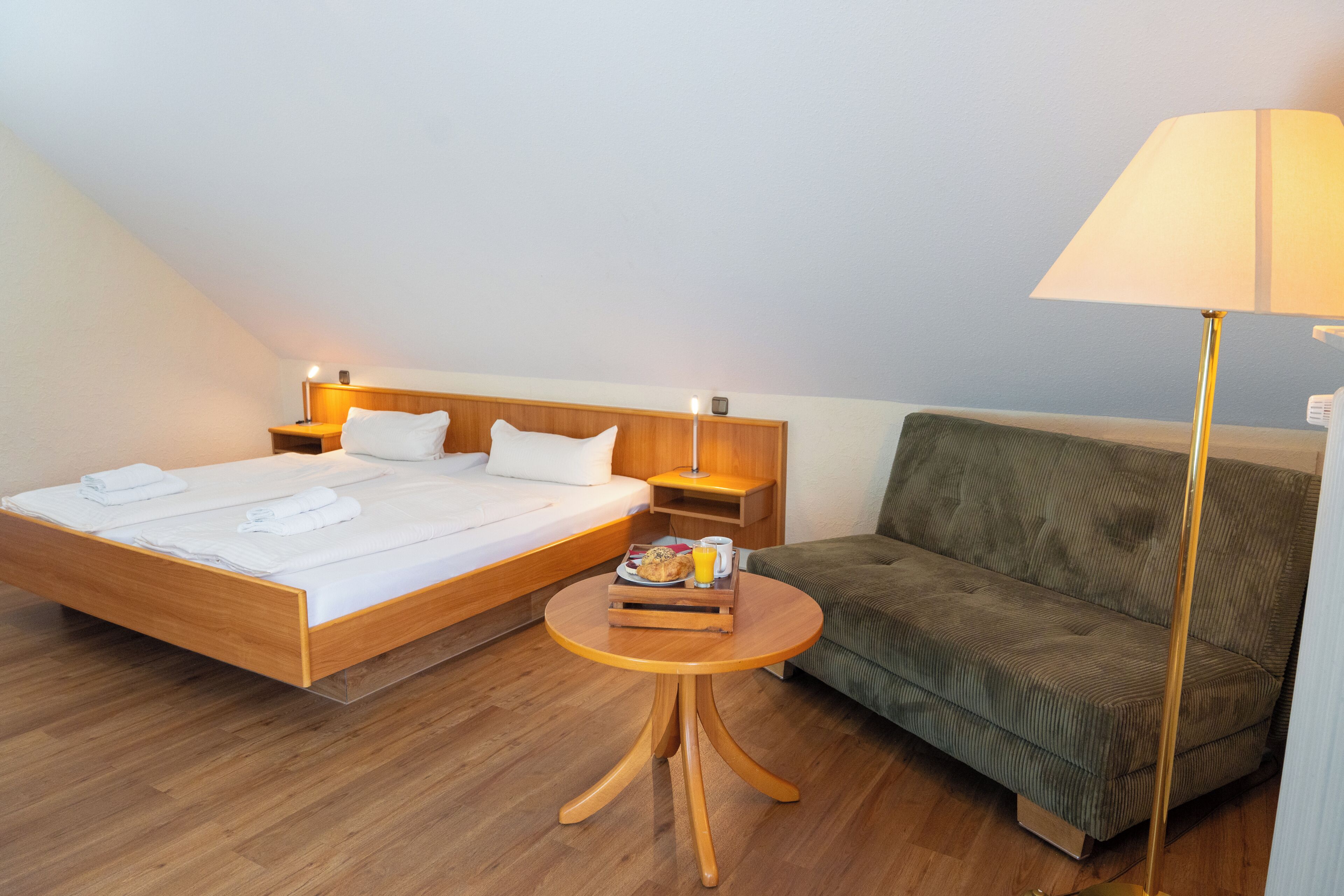 Quarto de hotel com cama, mesa de cabeceira, abajur, sofá e uma pequena mesa redonda com uma bandeja de itens de café da manhã.