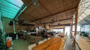 Ampla área de refeições interna com mesas e cadeiras de madeira, um bar e uma parede de pedra com teto de madeira.