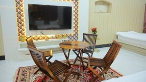 Uma sala de estar com televisão, cadeiras de madeira, uma mesa redonda e um tapete estampado.