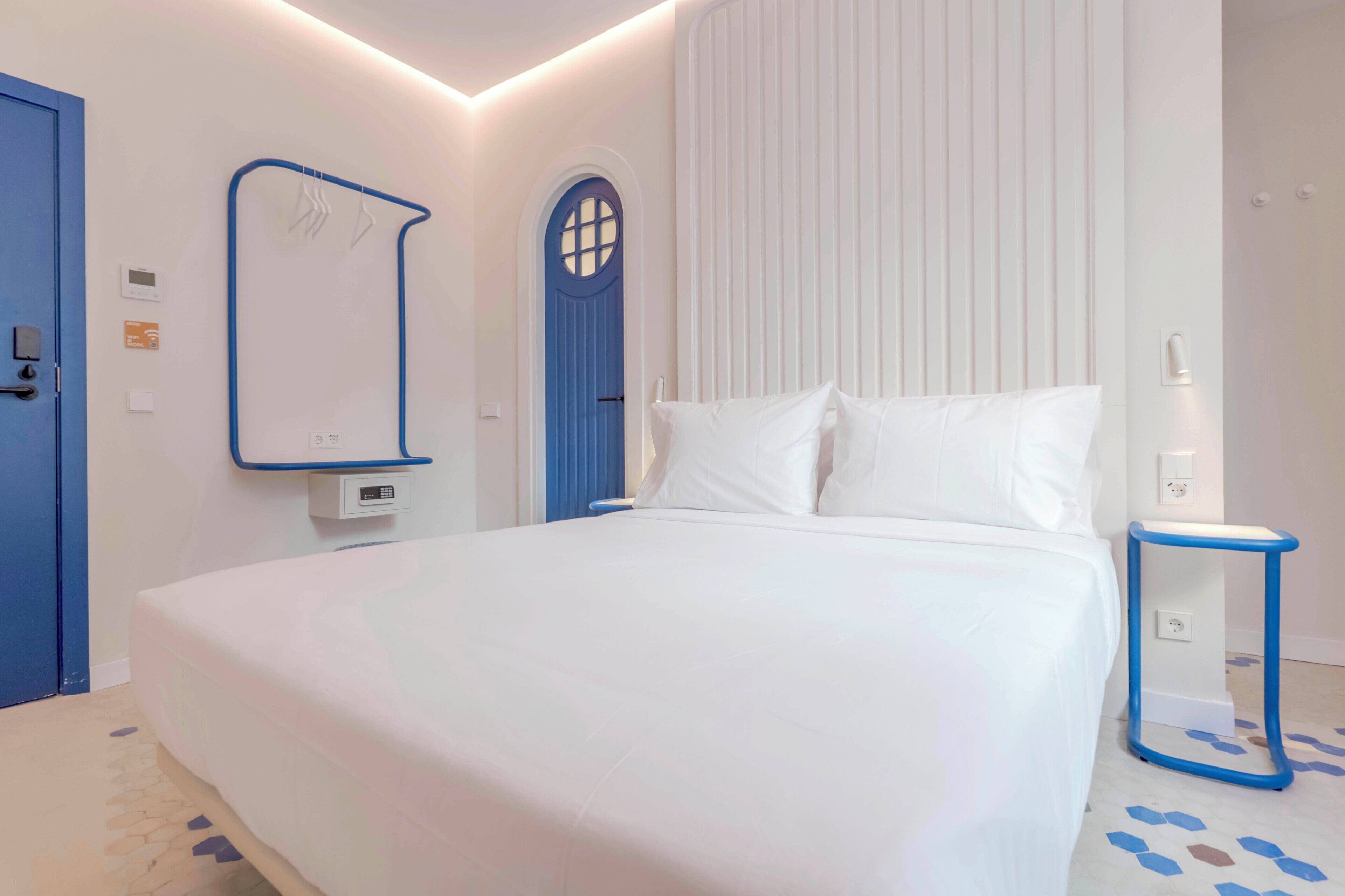 Quarto de hotel com uma cama, uma porta azul, uma parede branca e uma mesa de cabeceira.