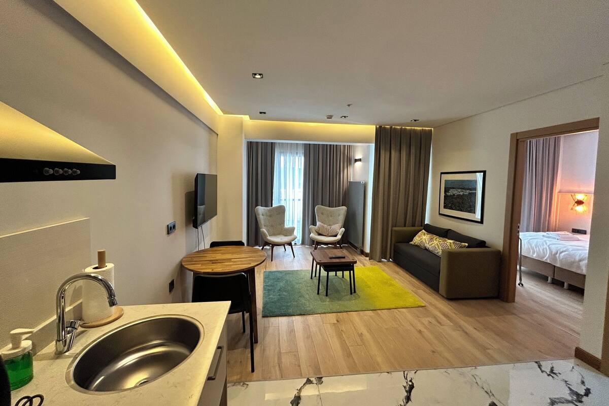 Quarto de hotel moderno com cozinha, área de estar com sofá e cadeiras, cama e uma janela grande com cortinas.