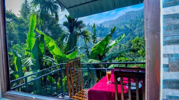 Uma varanda com mesa e cadeiras vermelhas, com vista para uma paisagem tropical exuberante com bananeiras e uma montanha ao fundo.