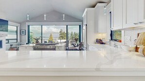 Cozinha moderna com uma ilha grande, armários brancos, iluminação pendente e vista para o exterior.