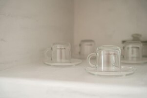 Uma bancada branca com três chávenas de vidro transparentes e pires, e um jarro de vidro transparente.