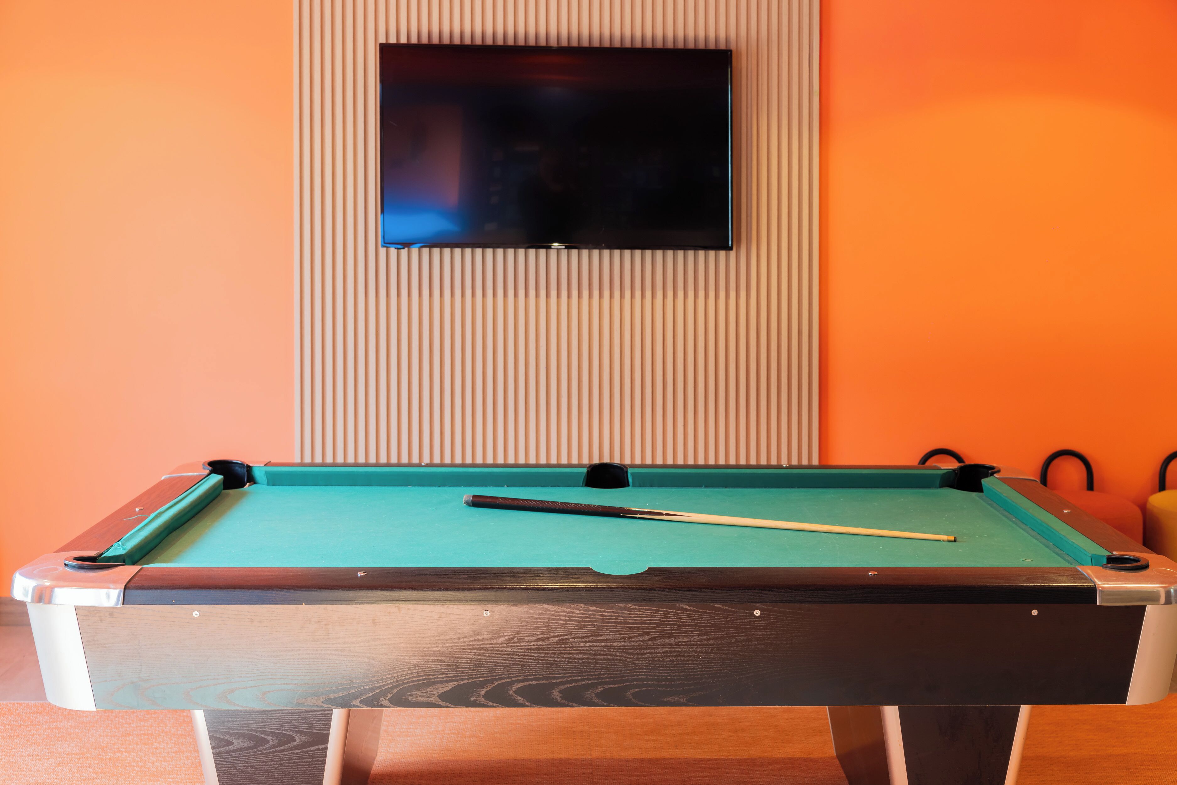 Uma mesa de bilhar com taco, uma televisão fixada na parede e uma parede listrada.