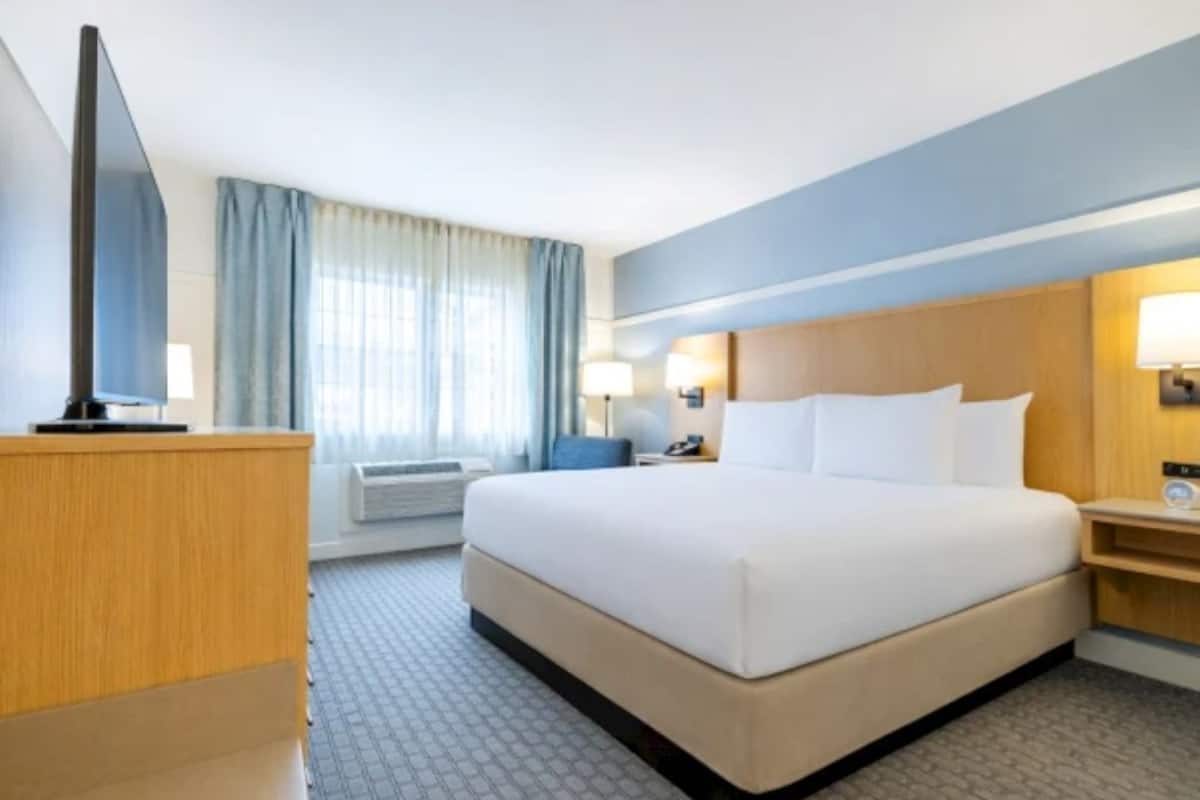 Quarto de hotel com uma cama grande, uma escrivaninha, uma televisão e uma janela com cortinas.