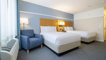 Quarto de hotel com duas camas, uma poltrona azul, um aquecedor e uma mesa de cabeceira com luminárias.