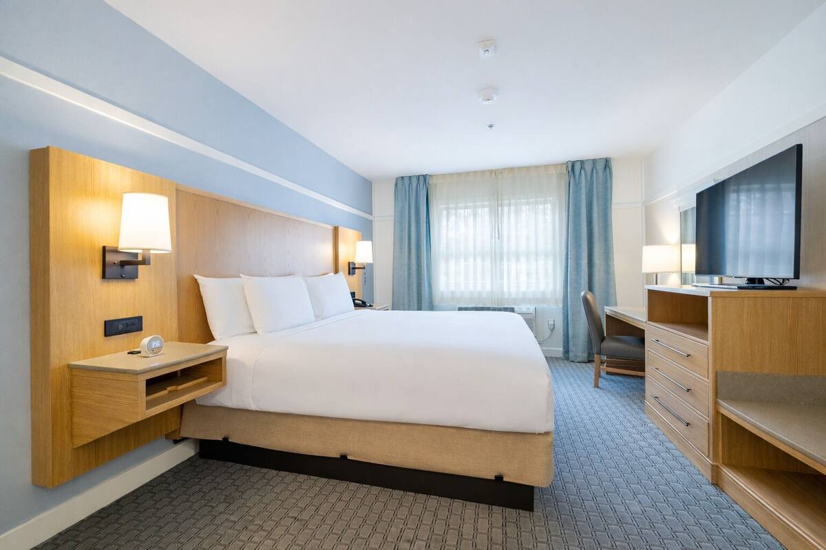 Quarto de hotel com uma cama grande, mesas de cabeceira, uma escrivaninha com cadeira, televisão e janela com cortinas.