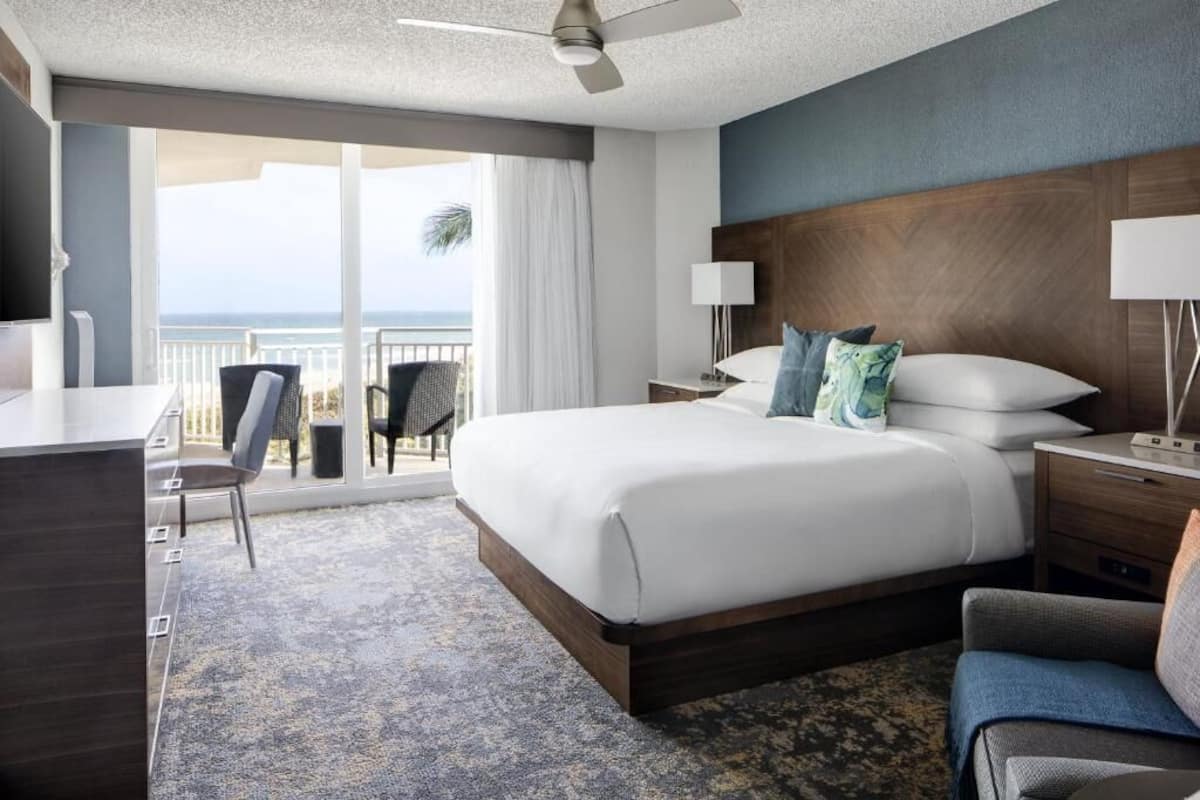 Quarto de hotel com uma cama grande, uma escrivaninha, uma cadeira, uma mesa de cabeceira e vista para o oceano através de grandes janelas.
