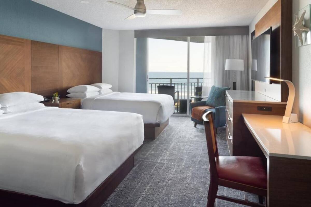 Quarto de hotel com duas camas, uma escrivaninha, uma cadeira, uma televisão e varanda com vista.
