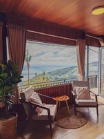 Quarto com teto de madeira, grande janela com vista para a paisagem, duas cadeiras, uma pequena mesa e um vaso com planta.