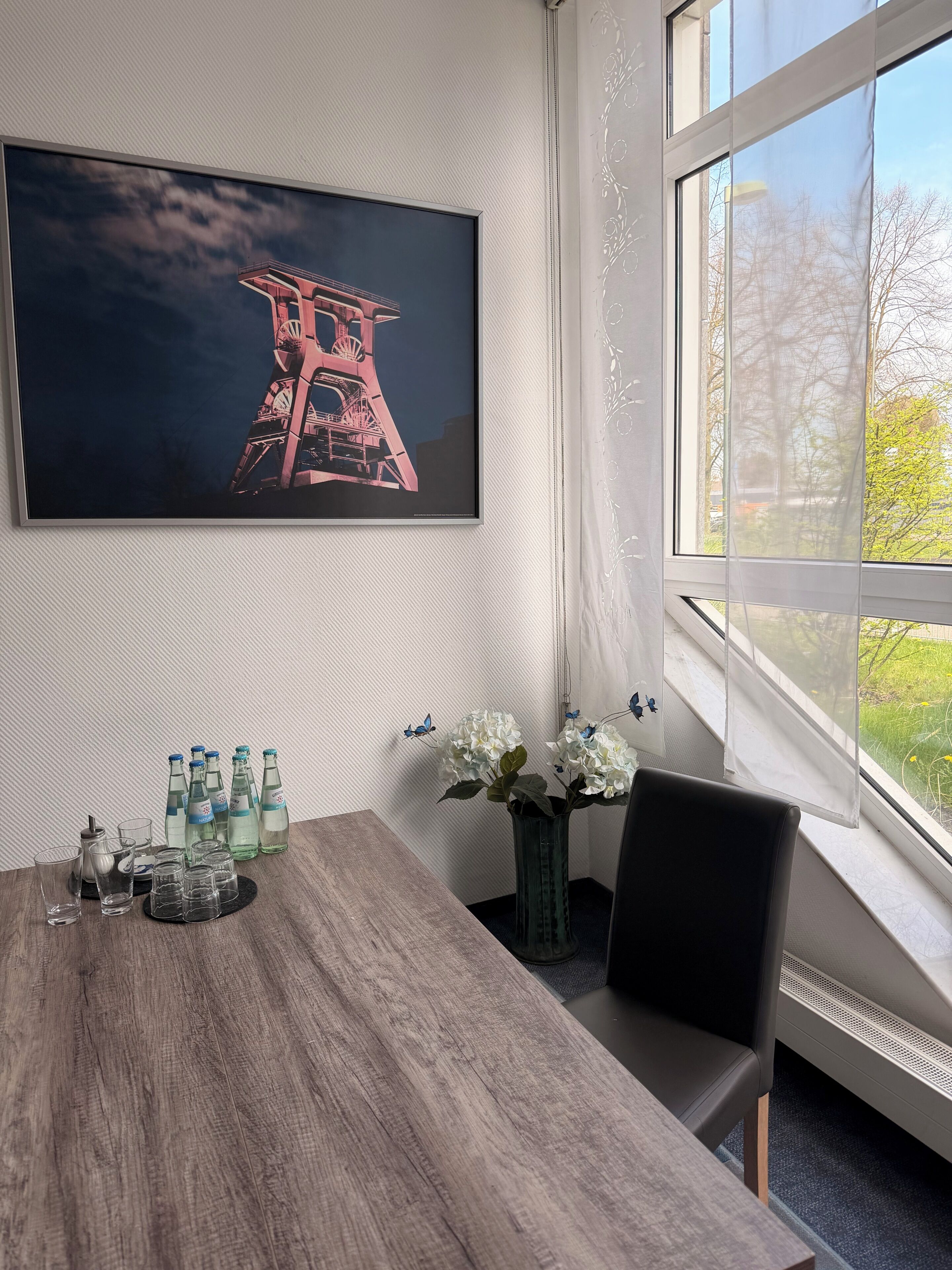 Quarto com mesa de madeira, um quadro emoldurado de uma torre, um vaso com flores e uma janela com vista para árvores.
