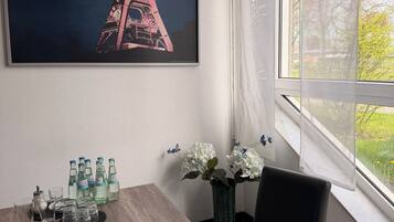 Quarto com mesa de madeira, um quadro emoldurado de uma torre, um vaso com flores e uma janela com vista para árvores.