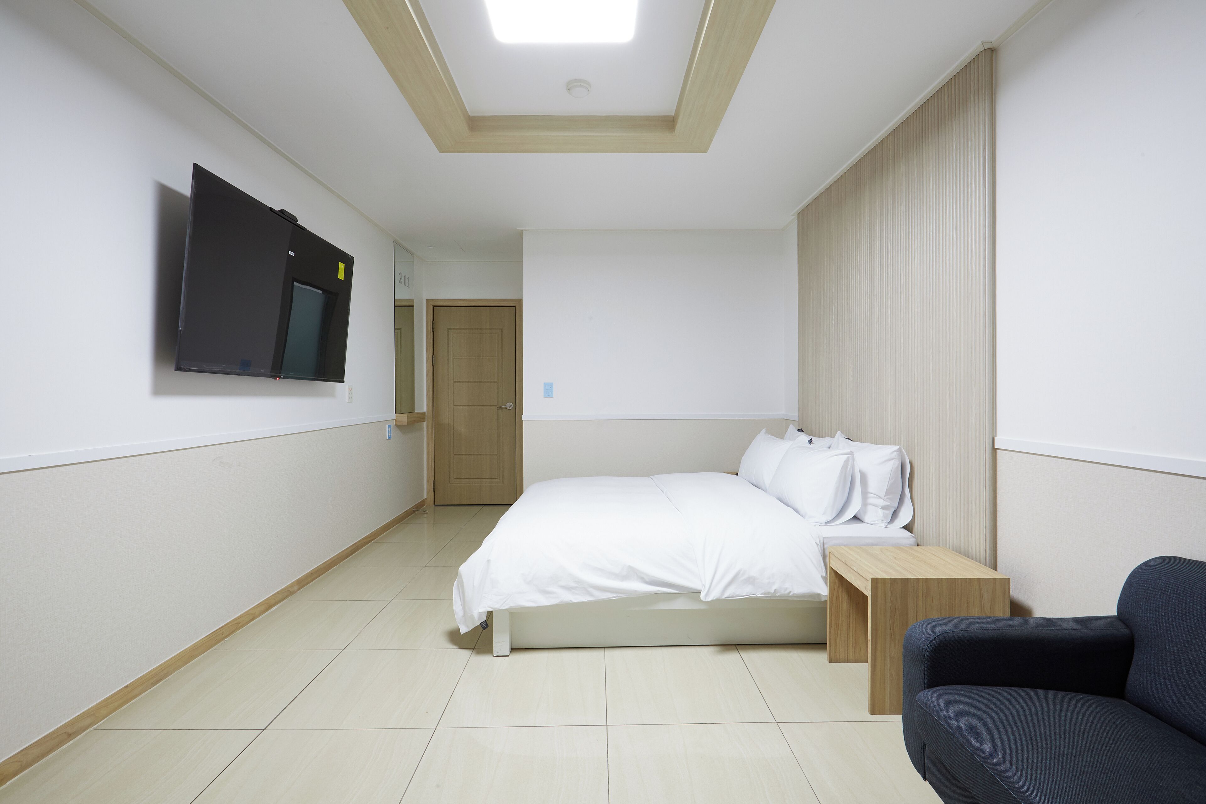 Quarto de hotel com cama, mesa de cabeceira, cadeira, televisão fixada na parede e porta para outro cômodo.