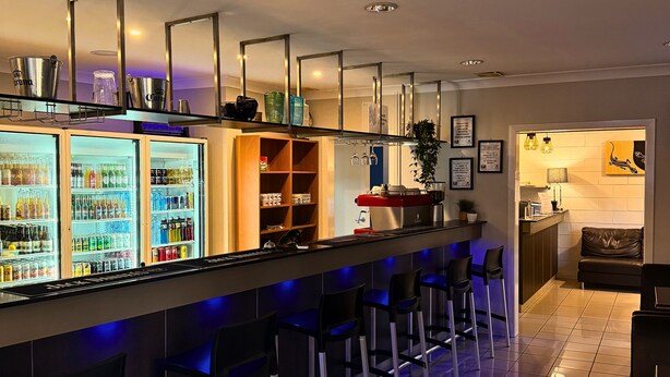 Uma área de bar com um balcão comprido, vários bancos e uma geladeira repleta de bebidas.