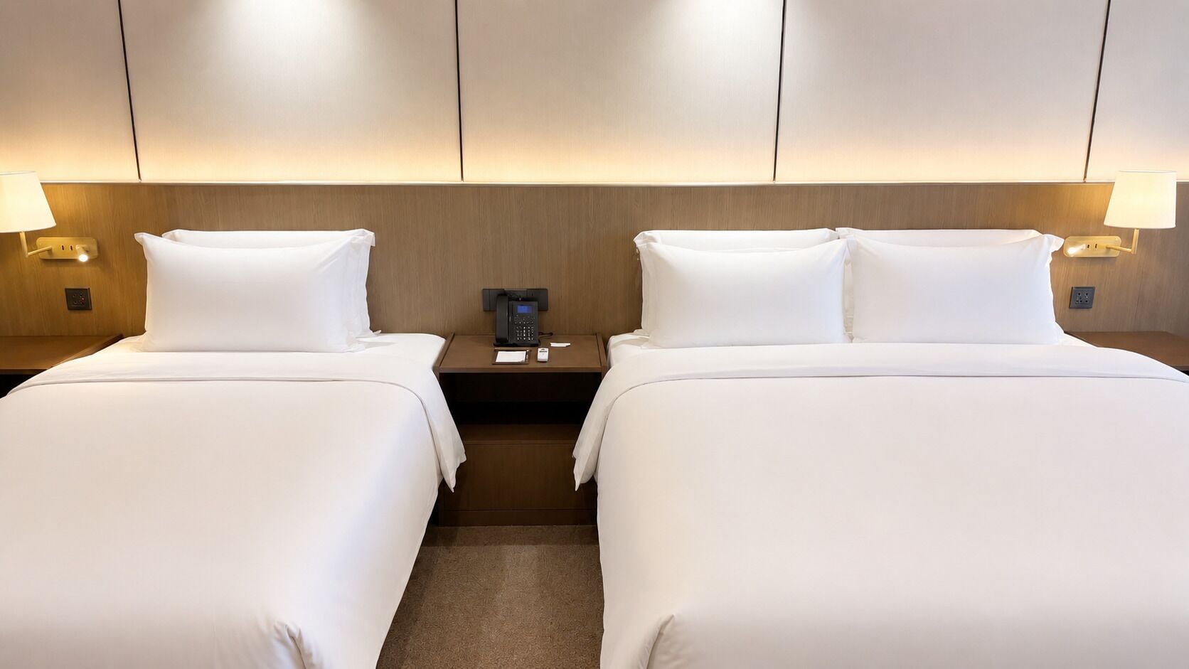 Quarto de hotel com duas camas, uma mesa de cabeceira com telefone e luminárias fixadas na parede.