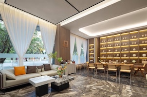 Um lobby de hotel moderno com um sofá, mesa de centro e cadeiras. Uma parede com prateleiras exibe objetos.