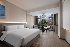 Quarto de hotel moderno com uma cama grande, área de estar com uma mesa, e vista da cidade através de amplas janelas.