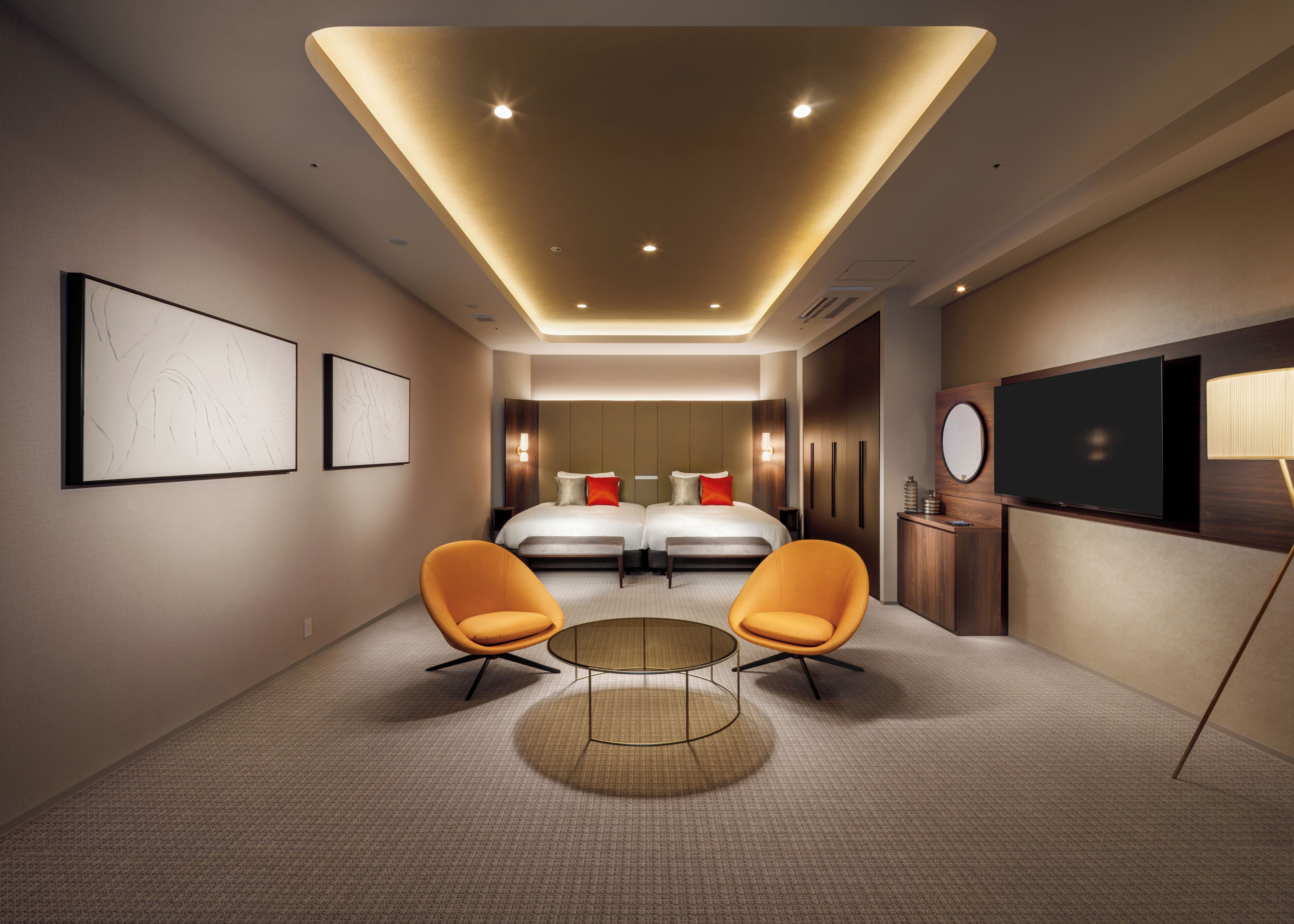 Quarto de hotel moderno com uma cama, duas poltronas laranja, uma televisão e obras de arte nas paredes.