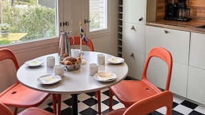 Uma área de refeições com uma mesa redonda, cadeiras laranja, uma cafeteira e vista para o verde lá fora.