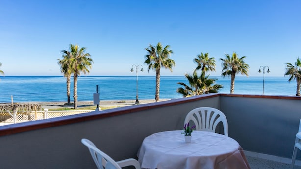 Varanda com mesa e cadeiras com vista para uma praia com palmeiras e o mar.