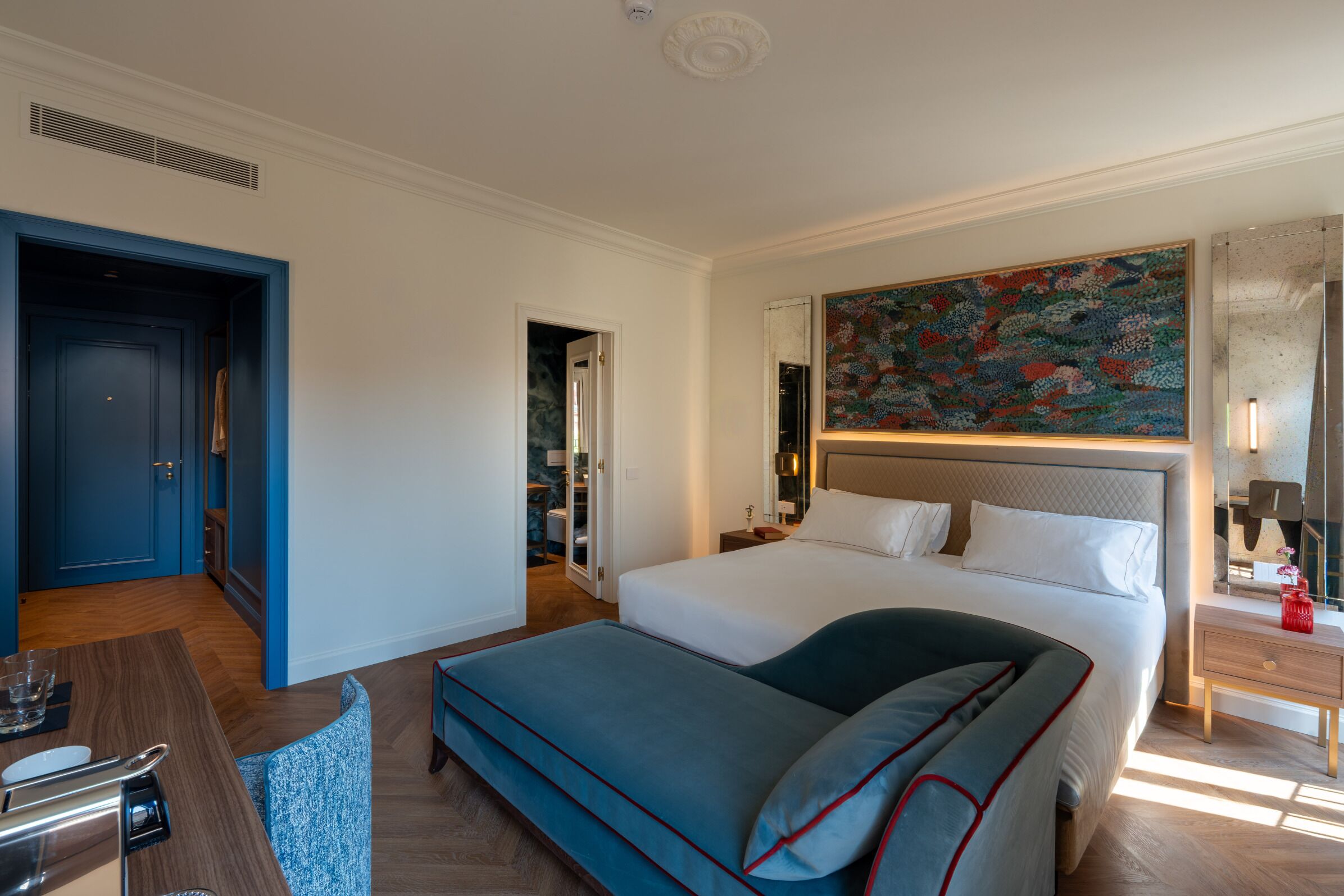 Quarto de hotel com uma cama grande, uma escrivaninha e uma cadeira. Há uma porta que dá para outro quarto e um quadro na parede.