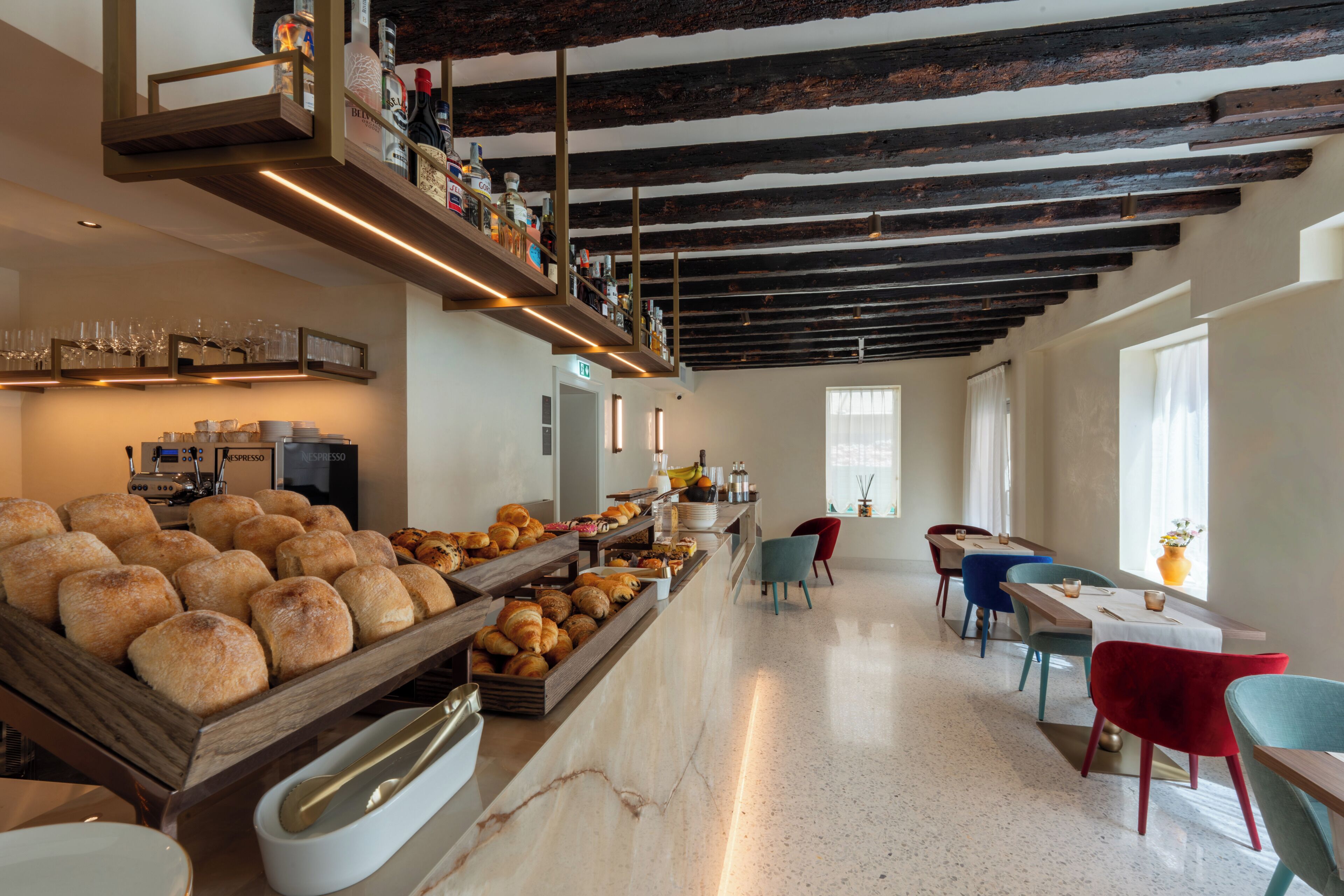 Um café com um balcão comprido exibindo pastéis e pão, uma máquina de café e várias garrafas em prateleiras.