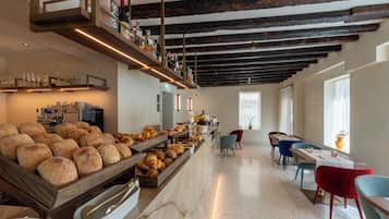 Um café com um balcão comprido exibindo pastéis e pão, uma máquina de café e várias garrafas em prateleiras.