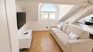 Um quarto moderno no estilo loft, com piso de madeira em padrão herringbone, um sofá branco, televisão na parede e janela tipo claraboia.