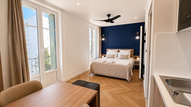 Quarto de hotel com cama, mesas de cabeceira, escrivaninha e uma cozinha compacta.