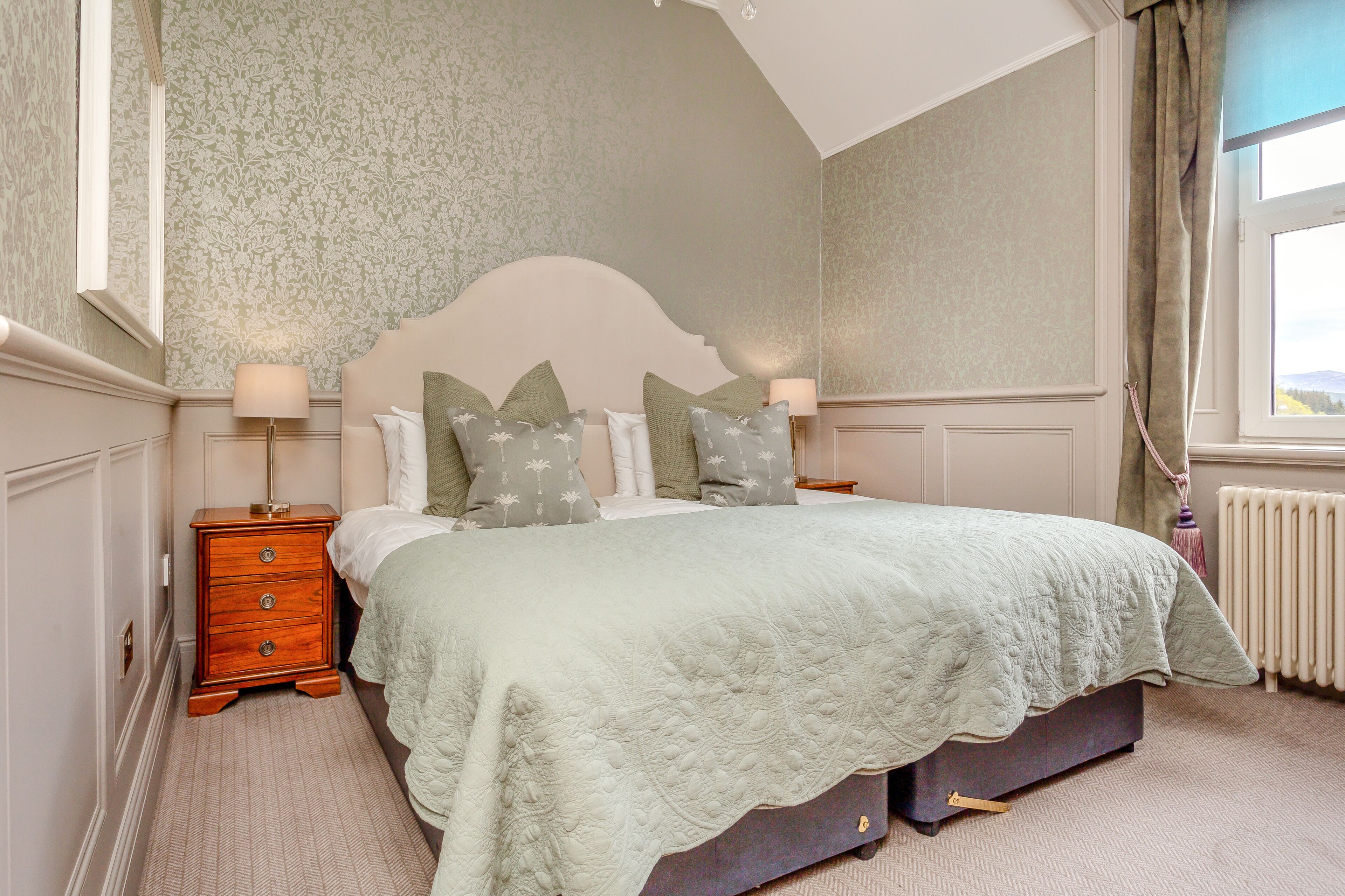 Quarto com cama, mesas de cabeceira, penteadeira e janela com cortinas.