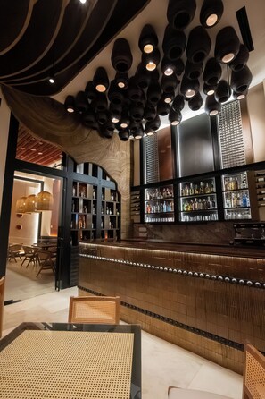Uma área de bar com balcão de madeira, prateleiras com garrafas e teto com luminárias circulares.