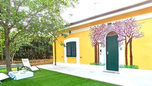 Um edifício com fachada amarela, porta verde e um mural de  árvores. Possui um gramado bem cuidado com duas cadeiras de descanso.