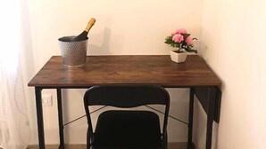 Uma pequena mesa de madeira escura com uma cadeira de metal na frente. Uma cesta de metal com uma garrafa e um vaso de planta estão sobre a mesa.