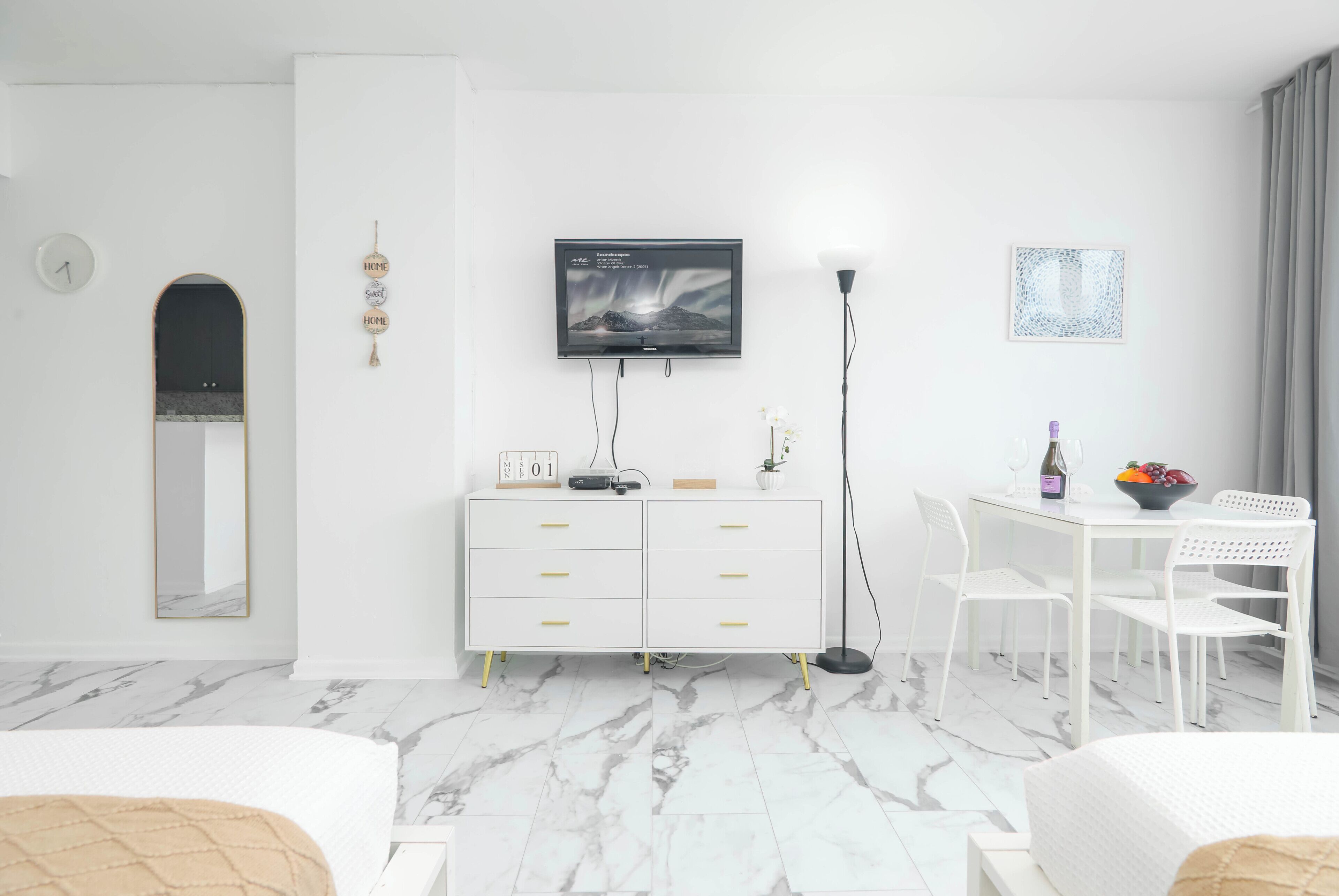 Quarto de hotel moderno com piso de mármore branco, TV de tela plana na parede, escrivaninha com cadeira e cama com manta bege.