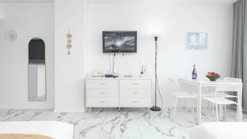 Quarto de hotel moderno com piso de mármore branco, TV de tela plana na parede, escrivaninha com cadeira e cama com manta bege.