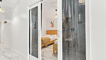 Quarto moderno com porta de vidro para um banheiro, cama de madeira e área de refeições com cadeiras.