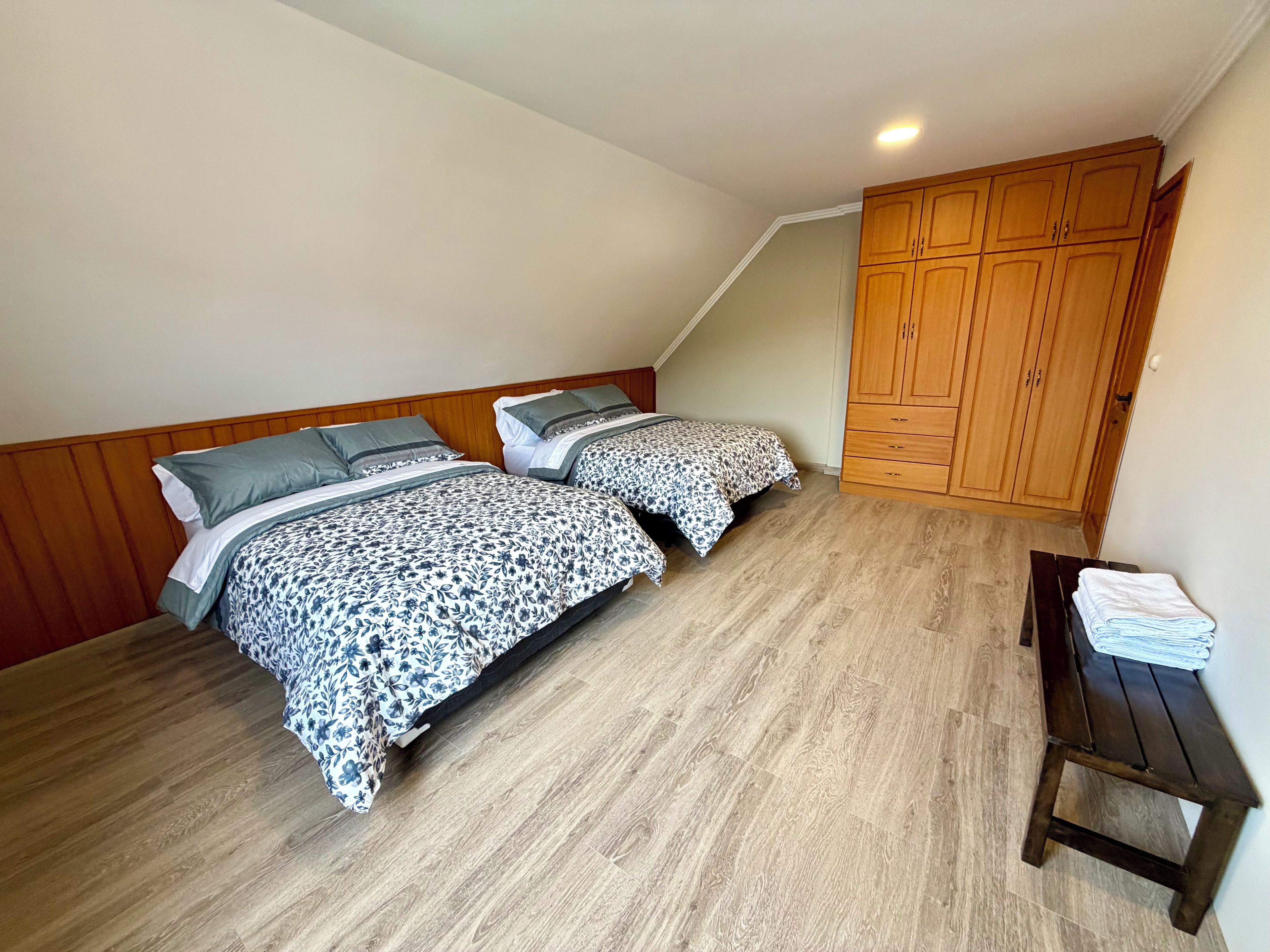 Quarto com duas camas, piso de madeira, guarda-roupa e um banquinho.