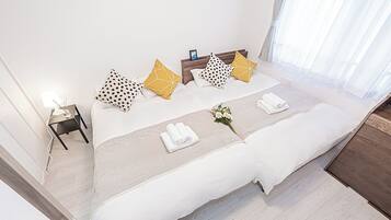 Cama bem arrumada com lençóis brancos, mesa de cabeceira com abajur e janela com cortinas leves.