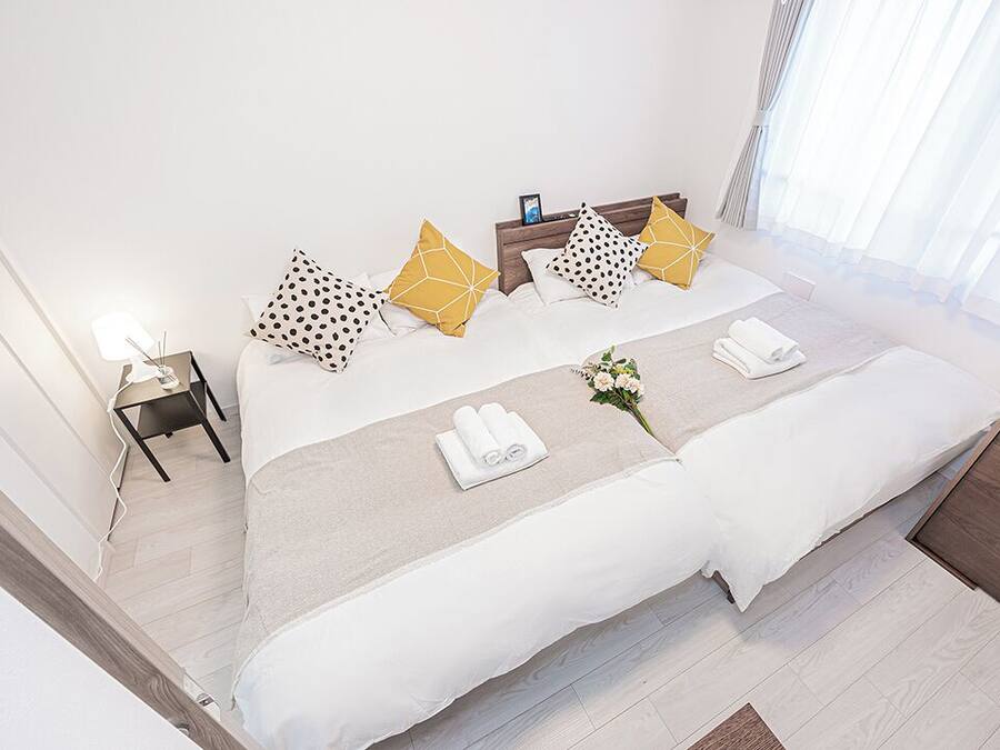 Cama bem arrumada com lençóis brancos, mesa de cabeceira com abajur e janela com cortinas leves.