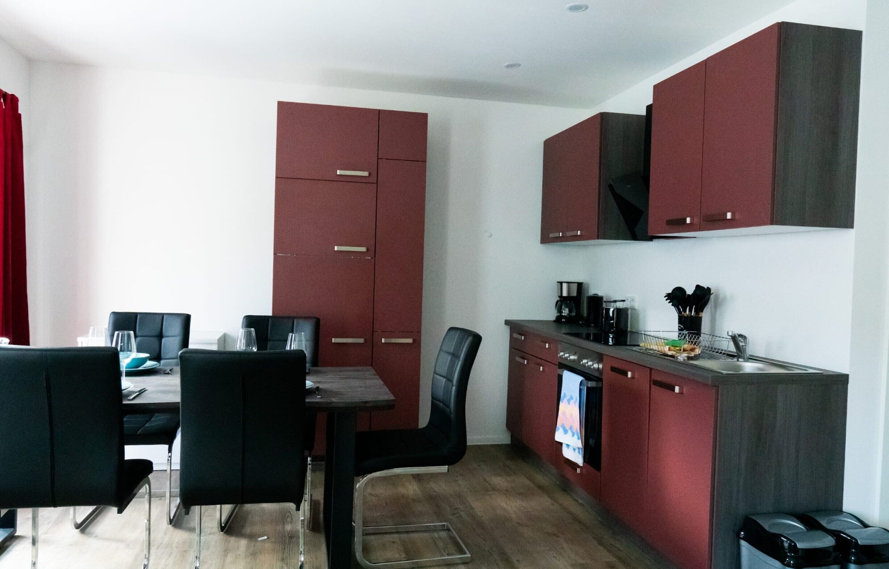 Cozinha compacta com armários vermelhos, área de refeições com cadeiras pretas e uma bancada pequena com cafeteira.